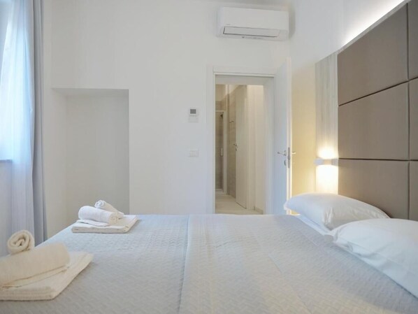 Quarto de hotel com cama, mesa de cabeceira, ar condicionado fixado na parede e porta que dá para outro cômodo.