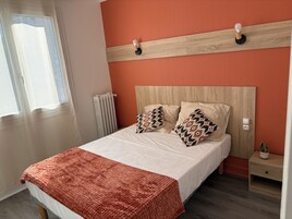 Cama bem arrumada com manta estampada, mesa de cabeceira com planta e luminária fixada na parede.