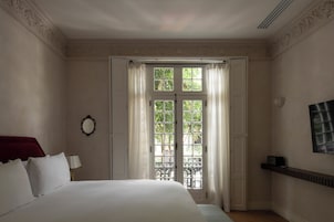 Quarto de hotel com cama, espelho, janela com cortinas e televisão presa à parede.