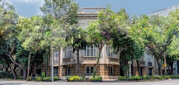 Um edifício histórico com grandes janelas e varanda, rodeado por vegetação exuberante.