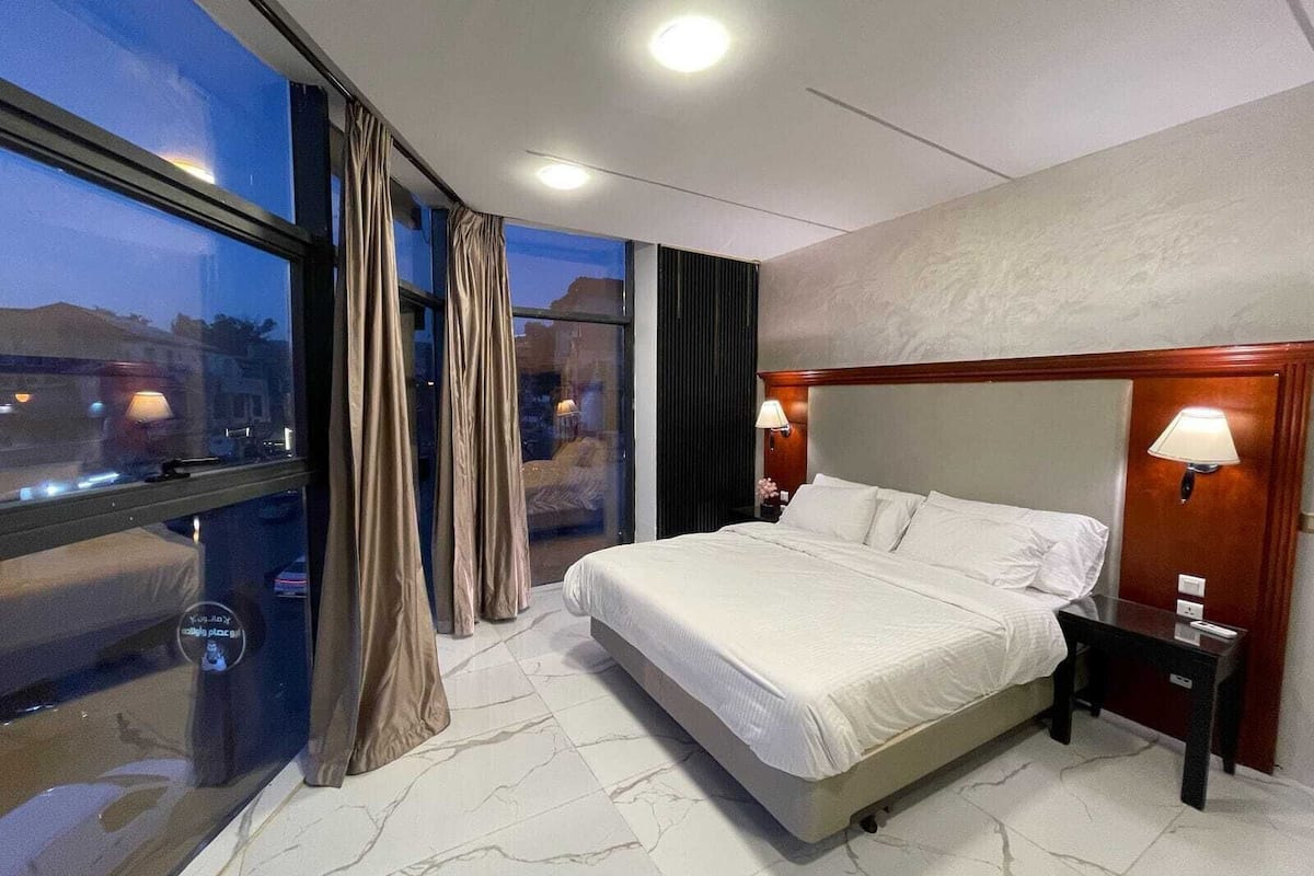 Quarto de hotel com uma cama grande, duas luminárias de cabeceira, vista pela janela e piso de mármore.
