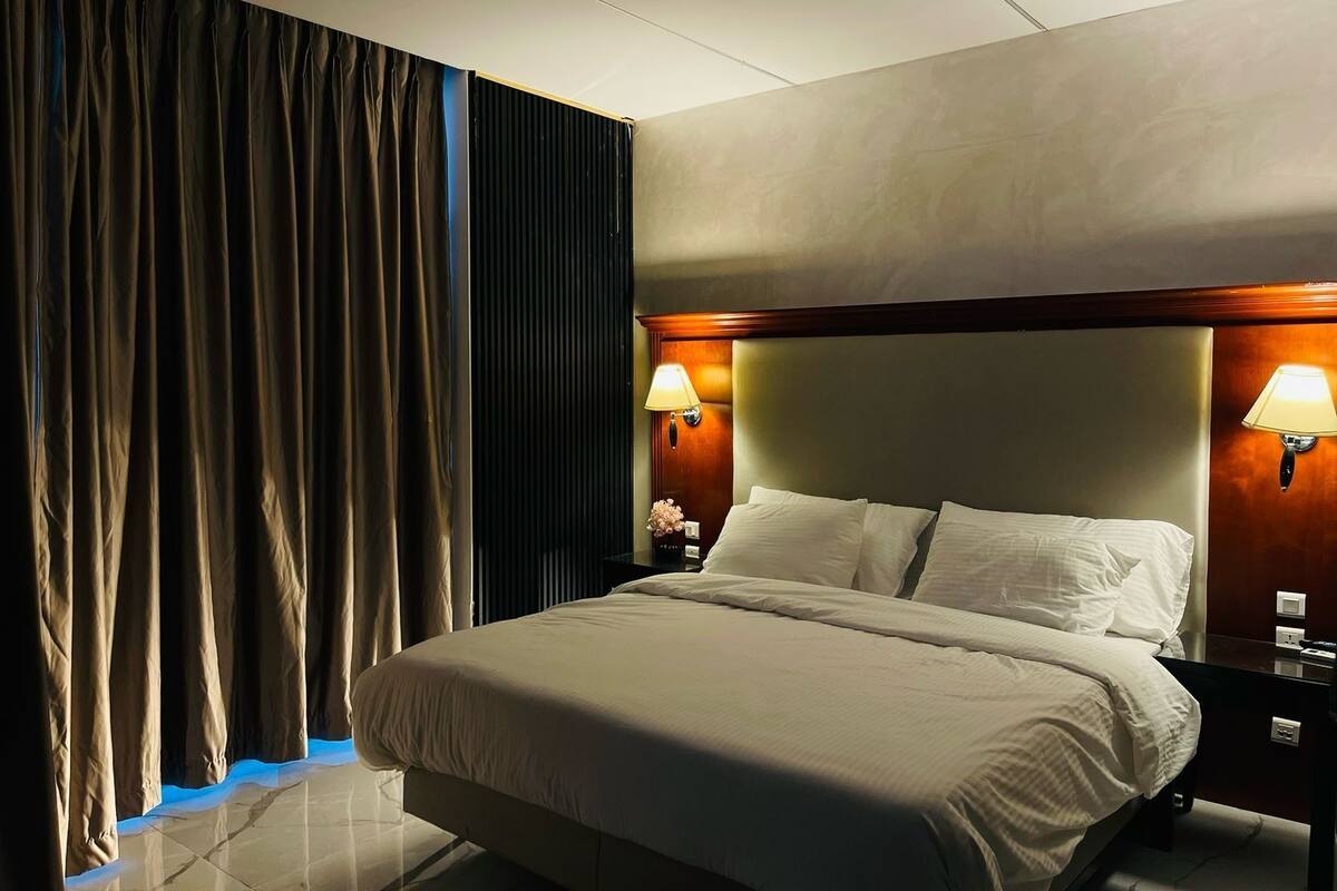 Quarto de hotel com uma cama grande, duas luminárias de cabeceira, uma cortina escura e uma mesa de cabeceira com um vaso de flores.