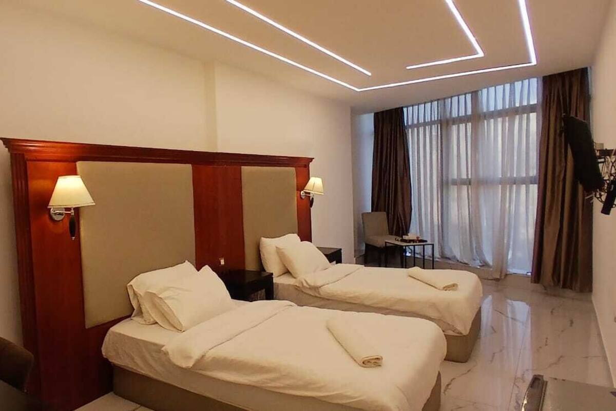 Quarto de hotel com duas camas, cabeceira de madeira, luminárias de cabeceira, uma mesinha e uma janela com cortinas.