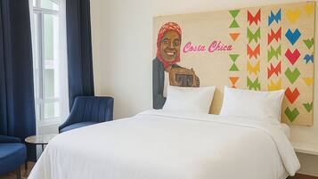 Quarto de hotel com uma cama grande, uma mesinha, uma poltrona azul e uma janela com cortinas.