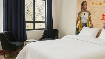 Quarto de hotel com uma cama grande, duas cadeiras, uma mesinha e uma janela com cortinas.