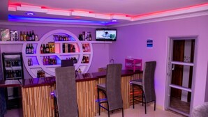 Uma área de bar com balcão de madeira, várias prateleiras com garrafas e banquetas.