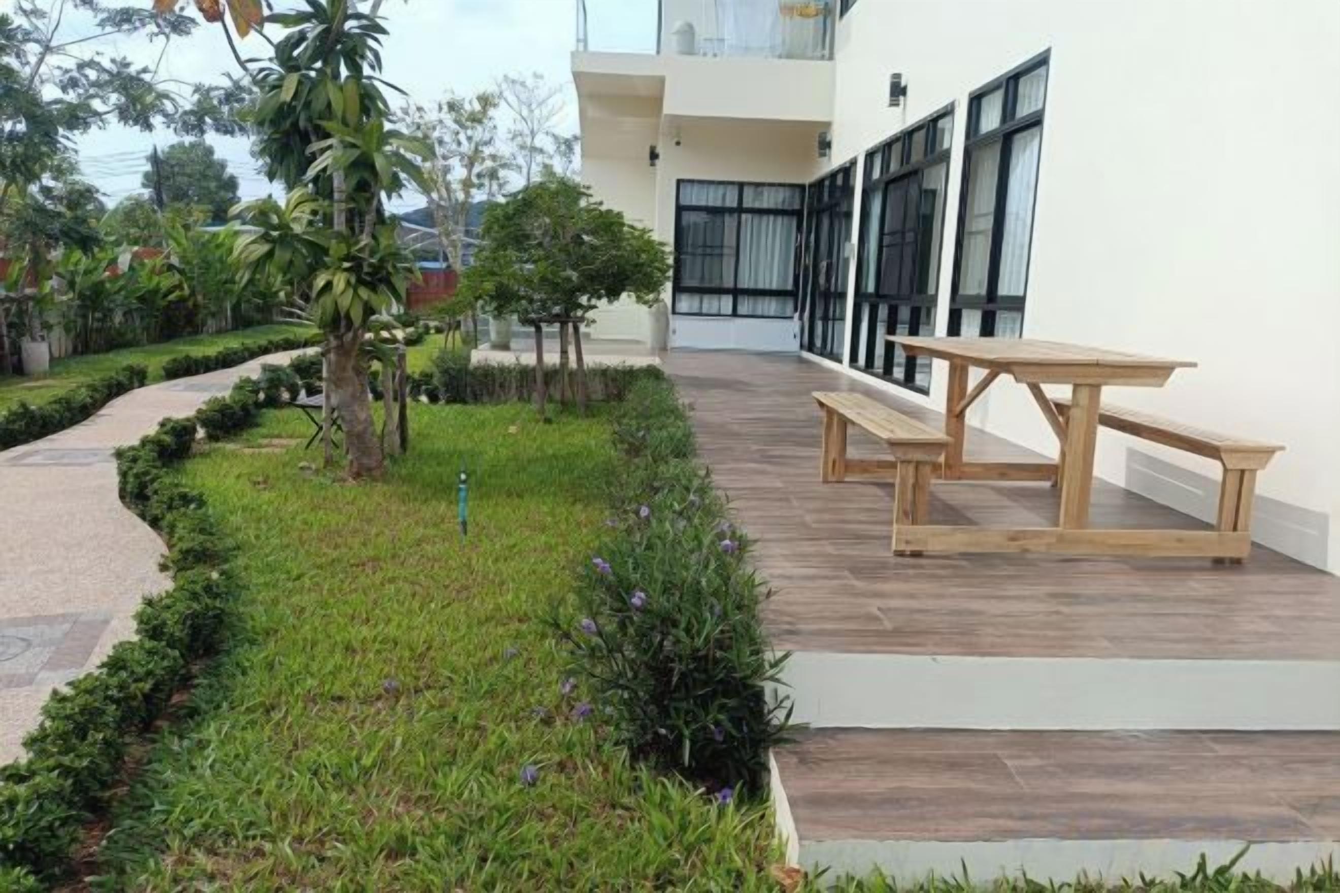 Um pátio externo com mesa e bancos de madeira, rodeado por um jardim bem cuidado com diversas plantas e um caminho pavimentado.