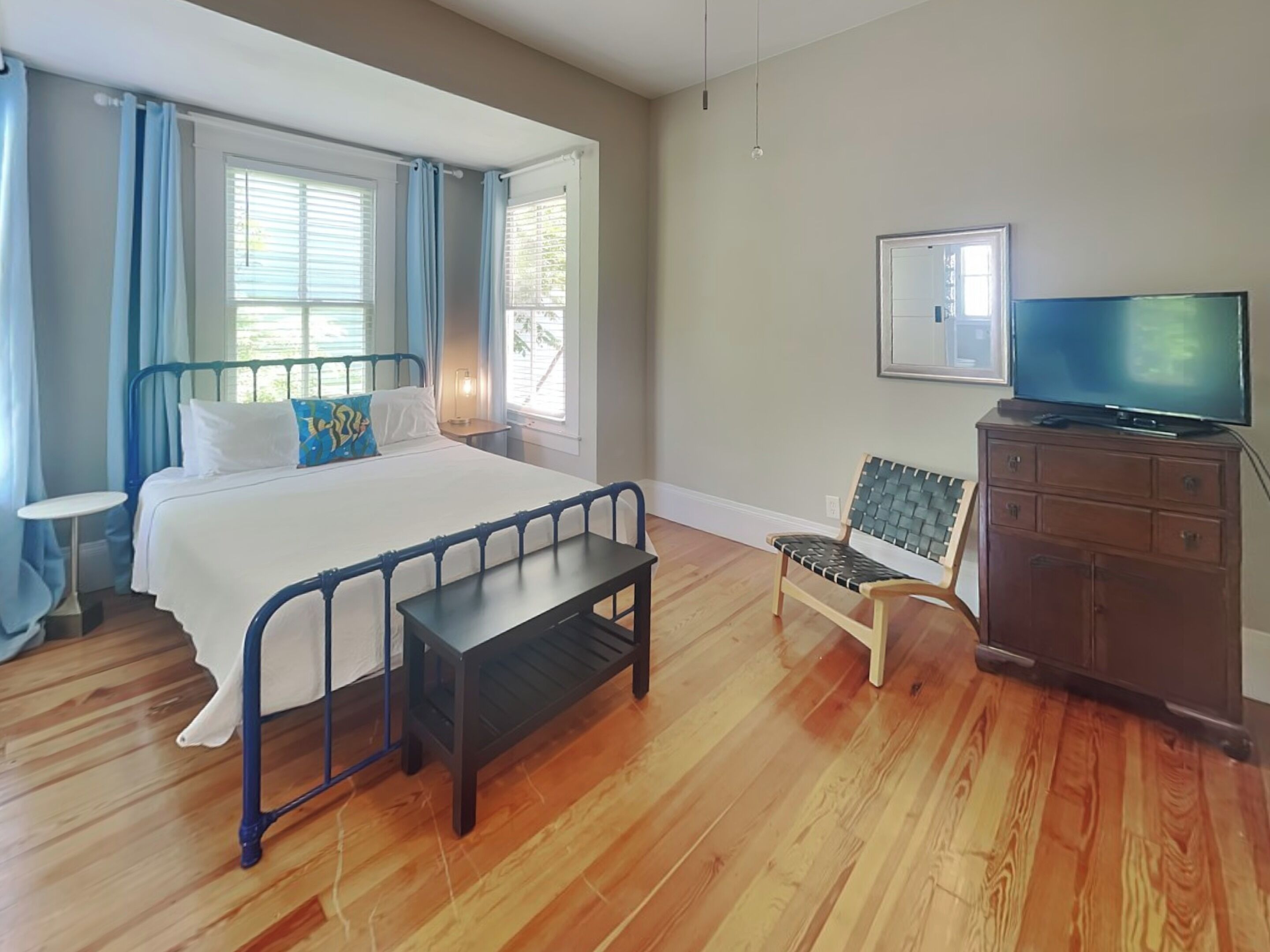 Um quarto com uma cama, um banquinho, uma cadeira, uma televisão e uma cômoda.