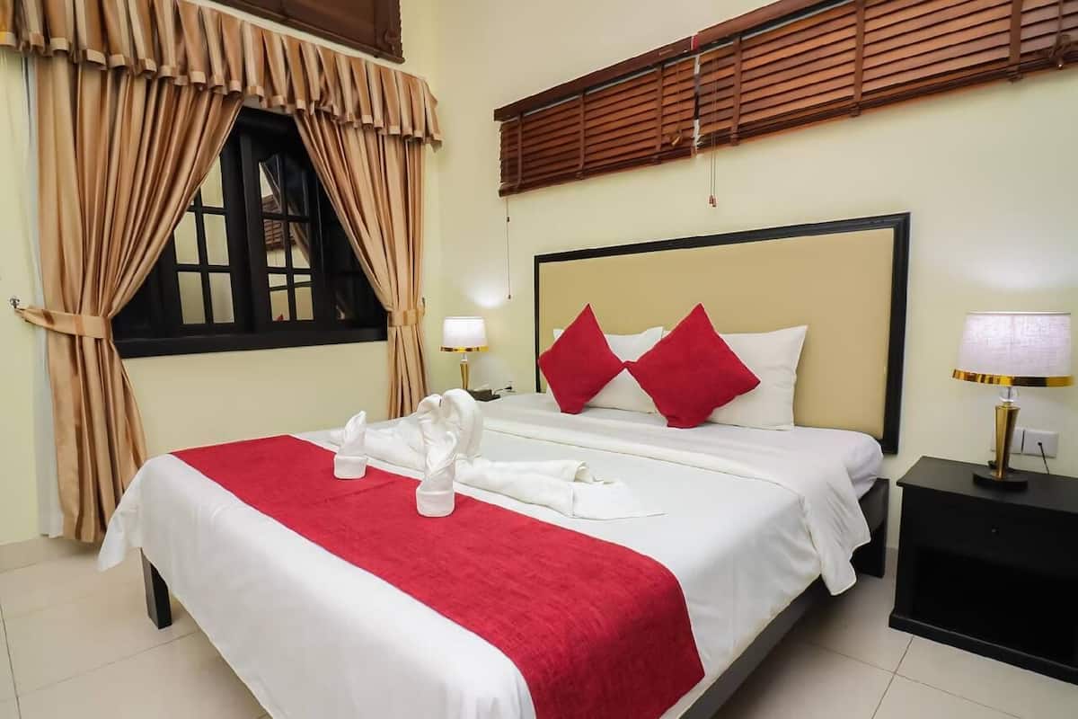 Quarto de hotel com cama, roupa de cama vermelha e branca, janela com cortinas, mesa de cabeceira com abajur e luminária na parede.