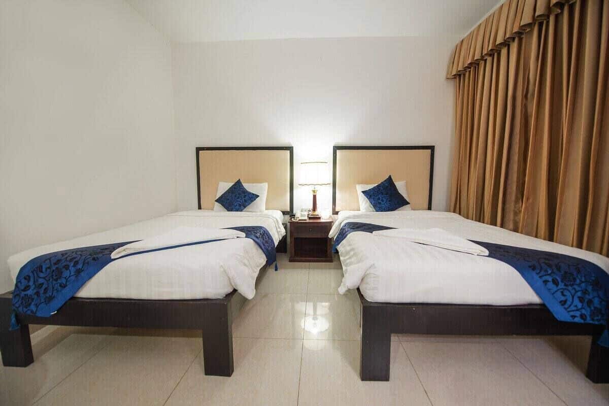Quarto de hotel com duas camas, uma mesinha de cabeceira entre elas, e uma cortina à direita.