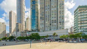 Uma área de praia com edifícios altos ao fundo, uma rede de voleibol e algumas pessoas.