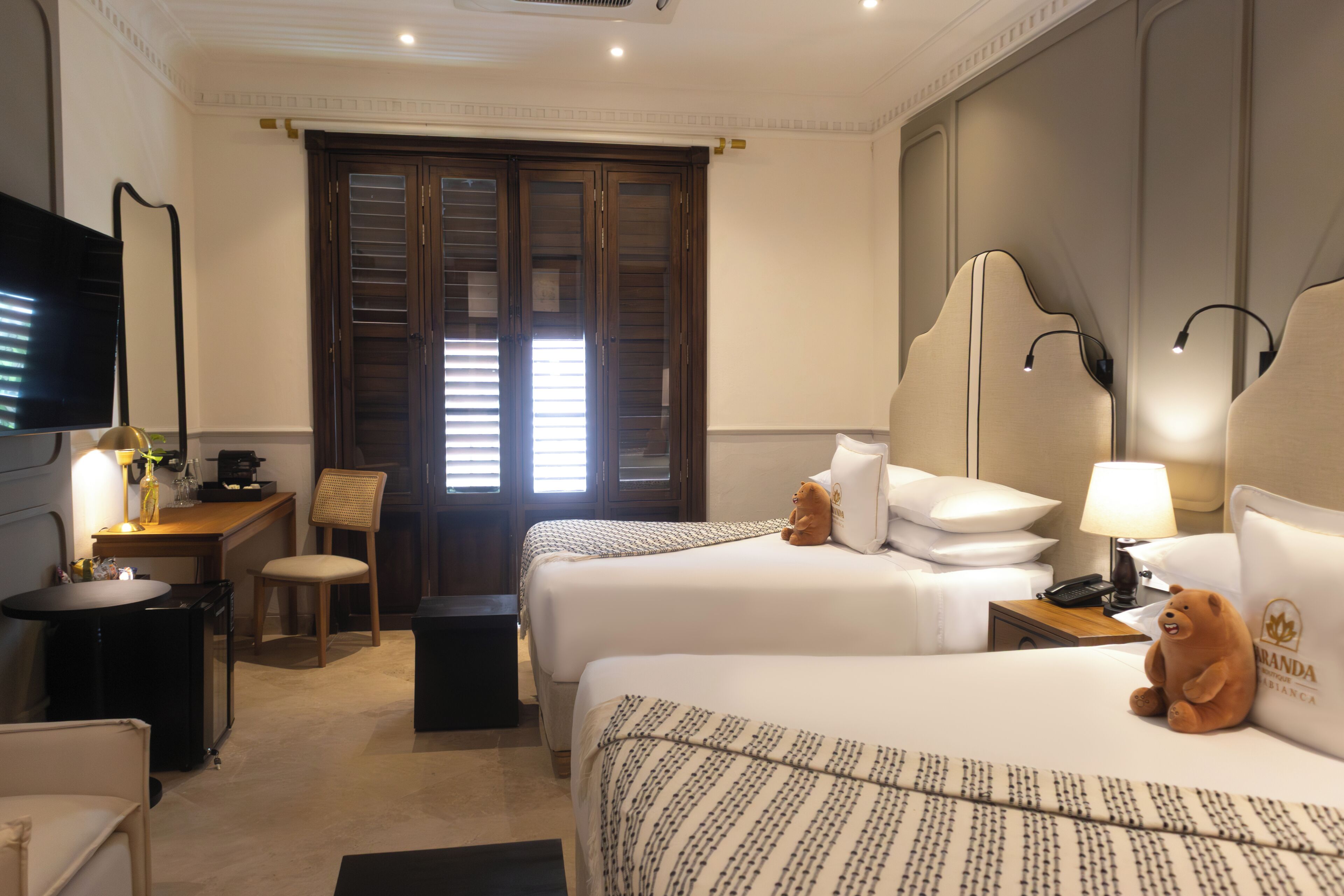 Quarto de hotel com duas camas, uma mesa de trabalho com cadeira, televisão e janela com persianas.