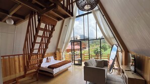 Quarto com cama de madeira, sofá, televisão e grande janela com vista para área verde e edifícios.