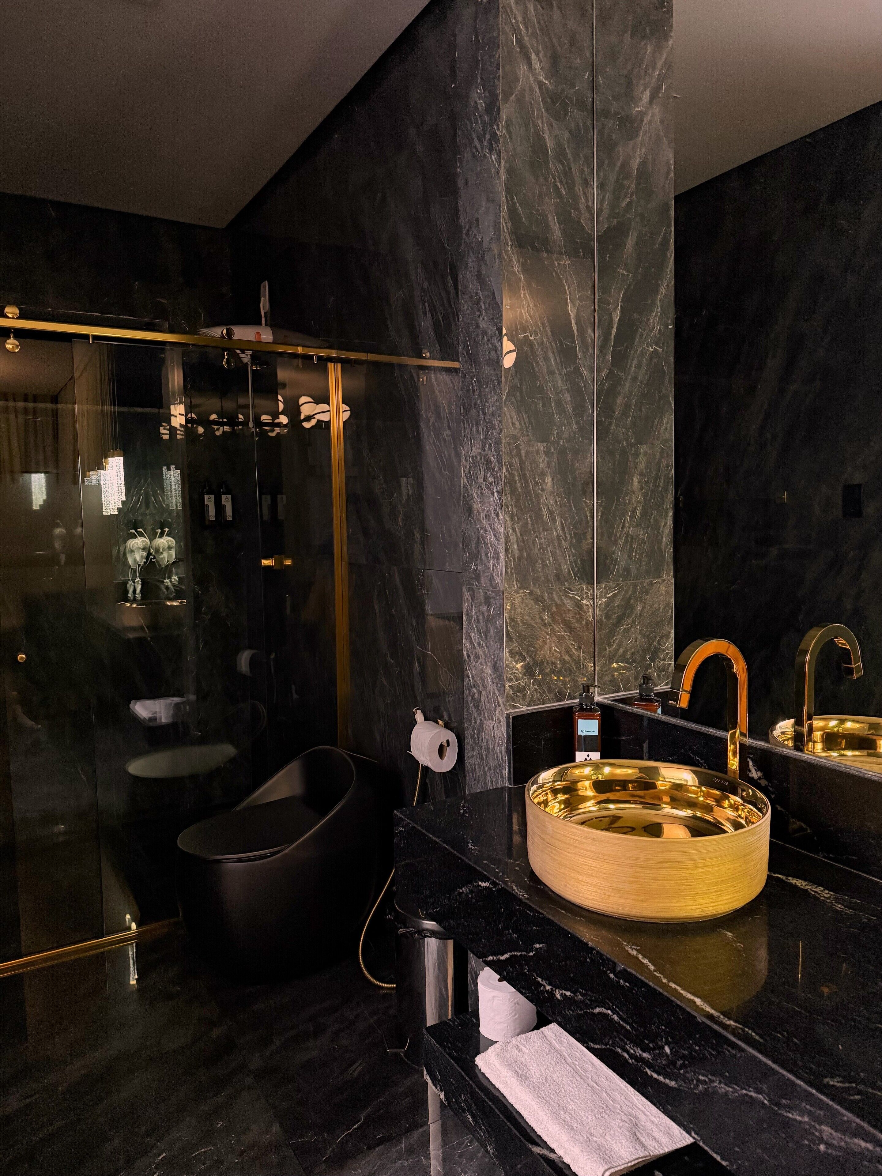 Uma casa de banho moderna com lavatório de mármore preto, torneiras douradas e zona de duche com divisória de vidro.
