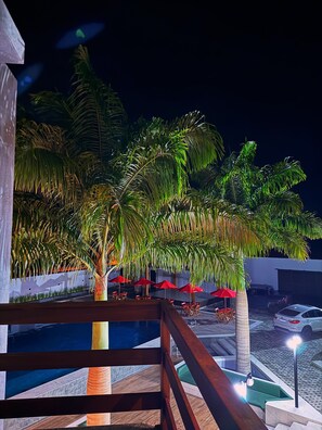 Vista noturna de uma área de piscina com palmeiras, guarda-sóis vermelhos e corrimão de deck de madeira.
