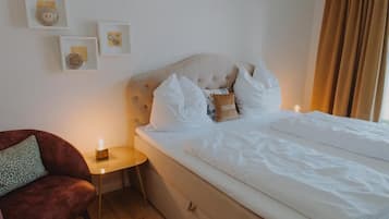 Uma cama bem arrumada com lençóis brancos, uma poltrona vermelha, uma pequena mesa de cabeceira com uma vela e obras de arte emolduradas na parede.