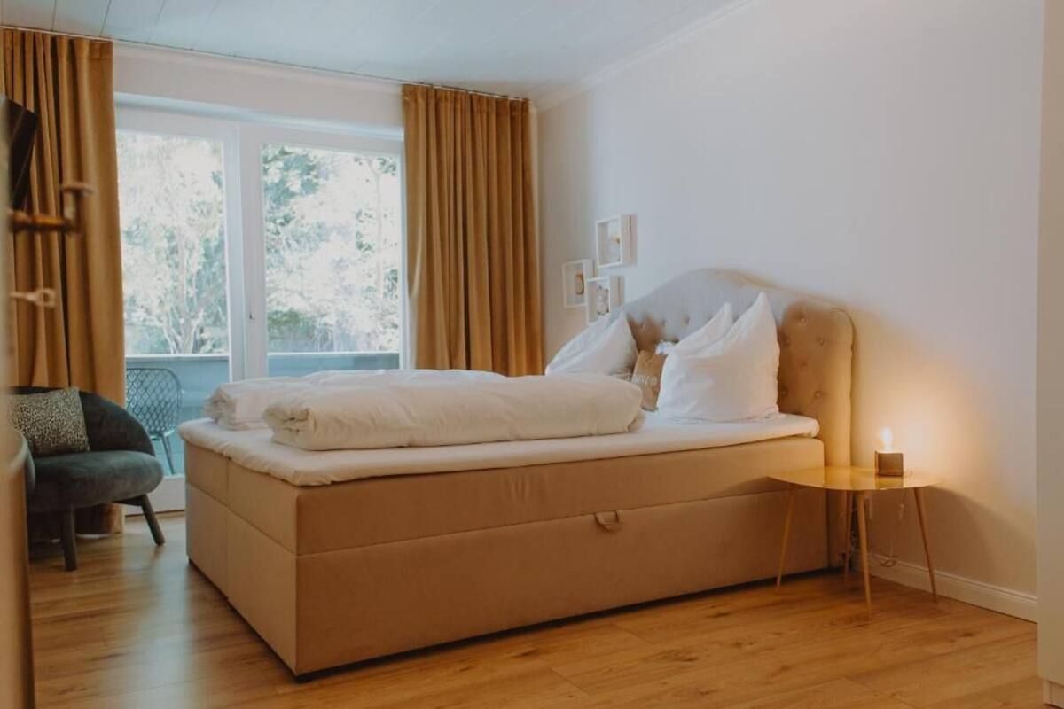 Quarto com cama, cadeira, mesa de cabeceira com abajur e janela com cortinas.
