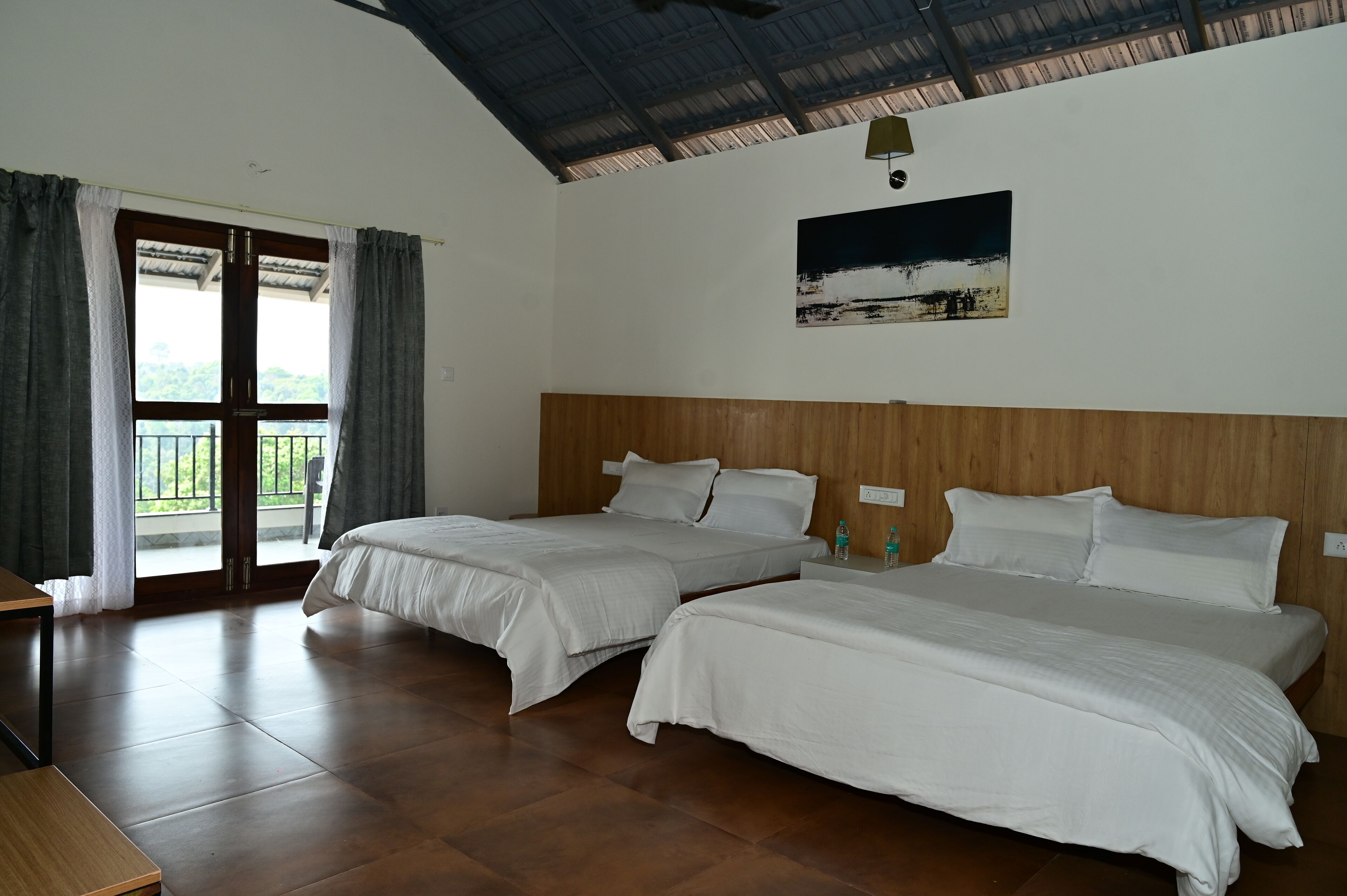 Quarto de hotel com duas camas, cabeceira de madeira, um quadro na parede e uma varanda com cortinas.