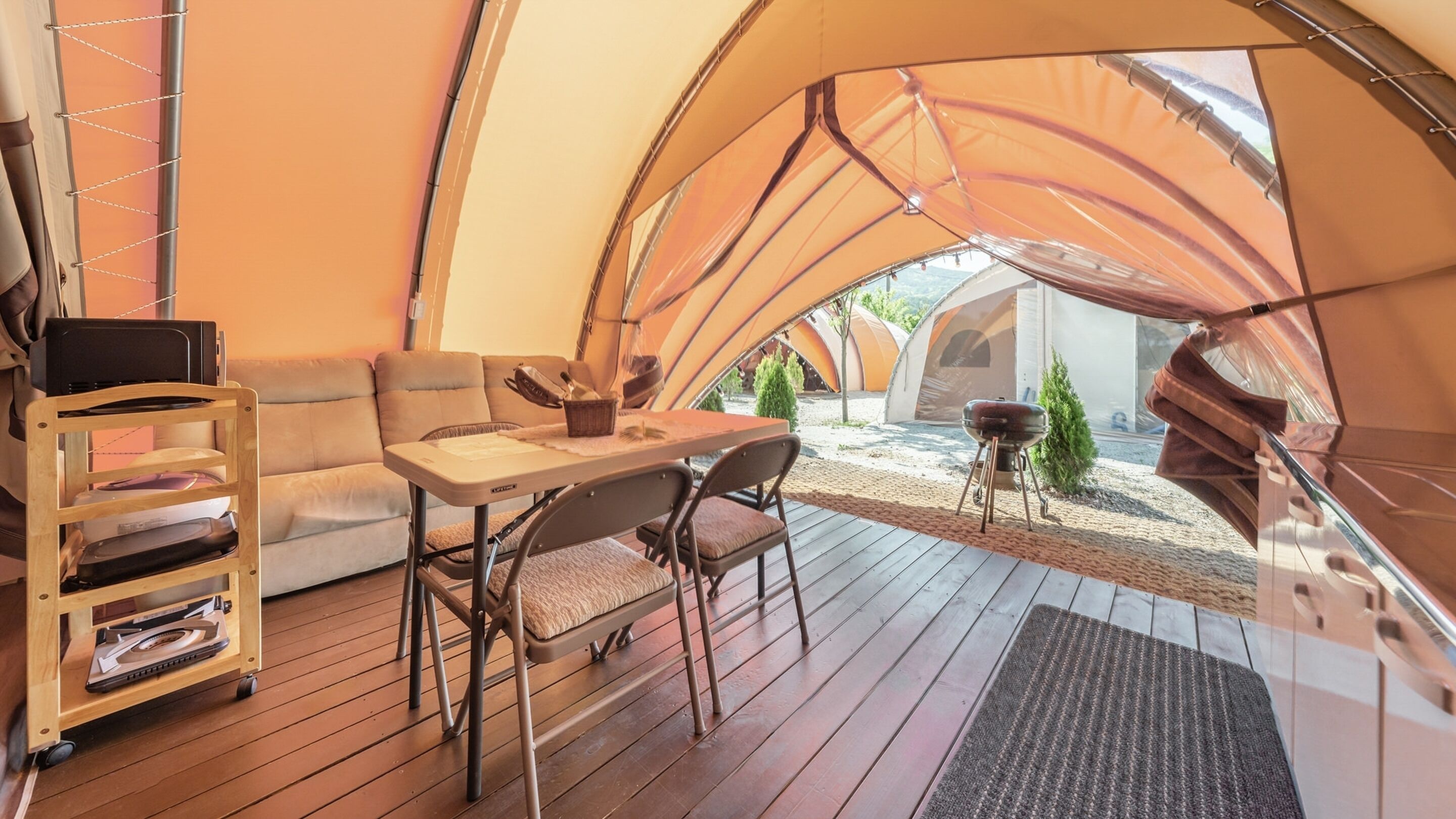 Interior acolhedor de uma tenda, com uma mesa de madeira, cadeiras, um sofá e um pequeno churrasco do lado de fora.