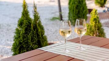 Dois copos de vinho branco sobre uma mesa de madeira com um jogo americano listrado, em ambiente externo com vegetação ao fundo.