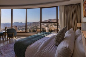Quarto de hotel com uma janela grande que oferece vista para a cidade, uma cama com almofadas estampadas, uma escrivaninha com cadeira e uma mesinha de centro com um vaso.