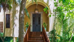 Escadaria que leva a uma porta com um candeeiro em cima, rodeada por vegetação exuberante e uma palmeira.