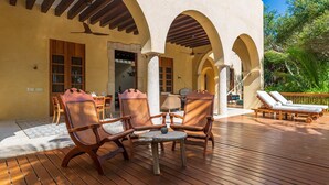 Um deck de madeira com móveis para área externa, incluindo cadeiras e uma mesa, sob uma cobertura com arcos e um ventilador de teto.