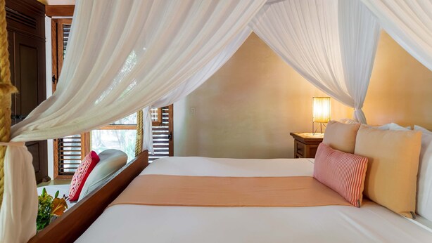 Quarto com cama de dossel, cabeceira de madeira, mesa de cabeceira com abajur e janela com persianas.