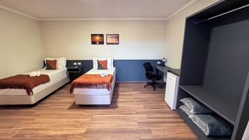 Quarto com duas camas de solteiro, uma escrivaninha com cadeira, um frigorífico pequeno e uma televisão.