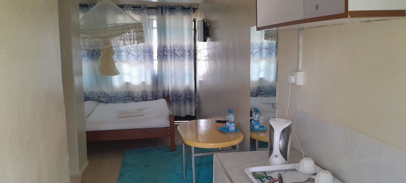 Quarto pequeno e compacto com uma cama de solteiro, uma mesinha com uma cadeira, um espelho e uma televisão na parede.
