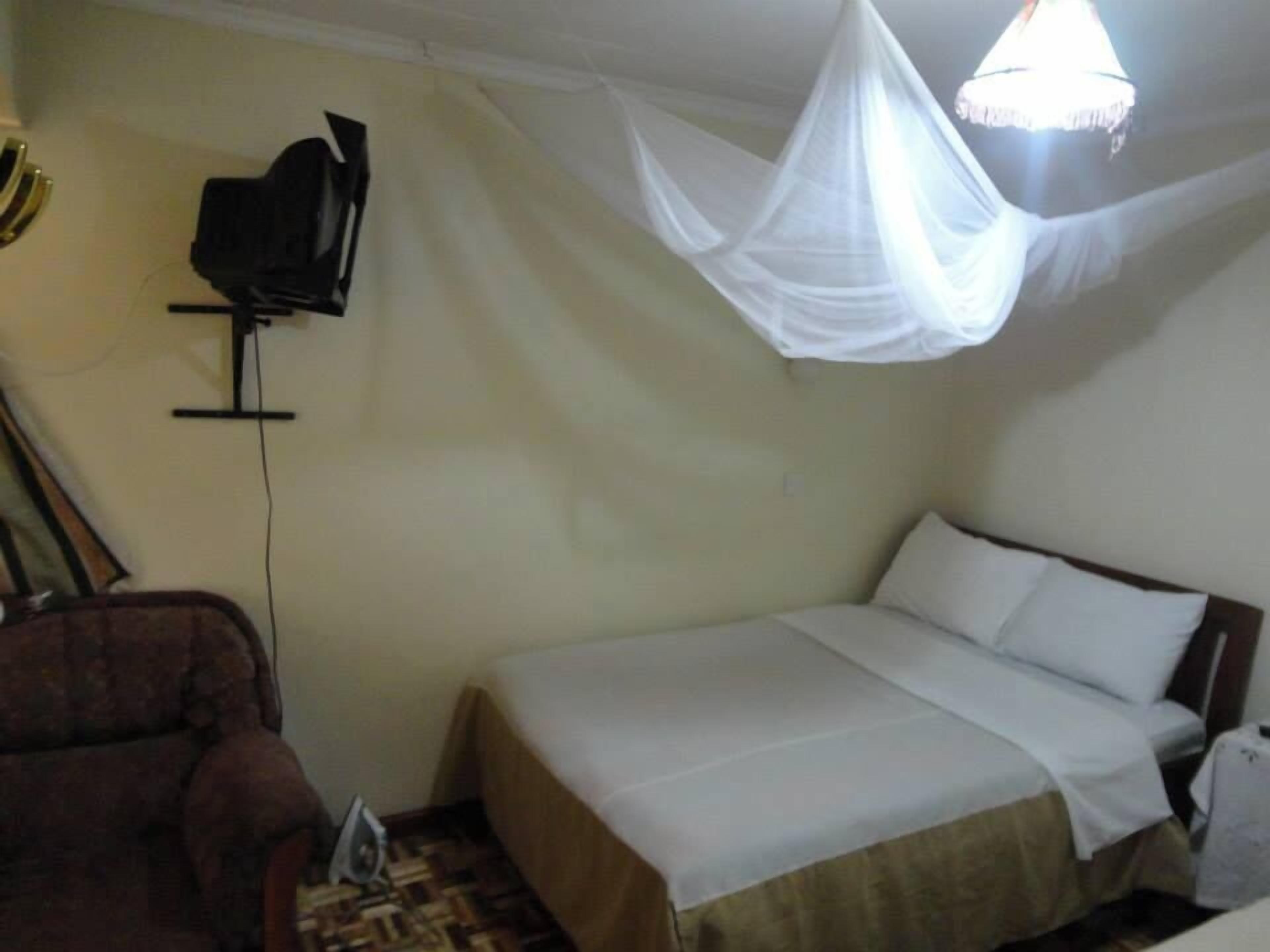 Uma cama de solteiro com roupa de cama branca, uma televisão presa na parede e uma mosquiteira pendurada sobre a cama.