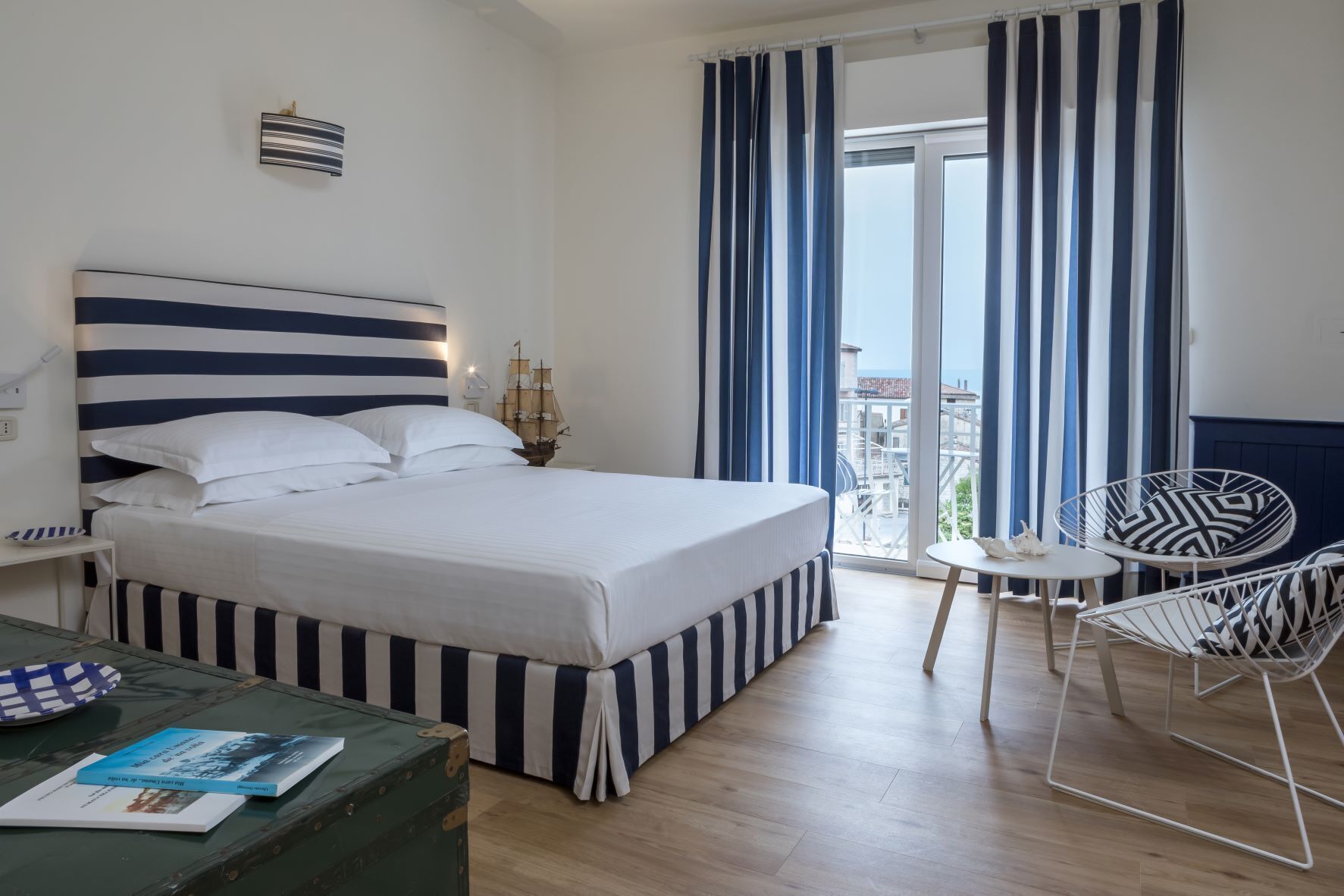 Quarto de hotel com uma cama, uma mesinha, uma cadeira, uma mala e vista para o exterior.