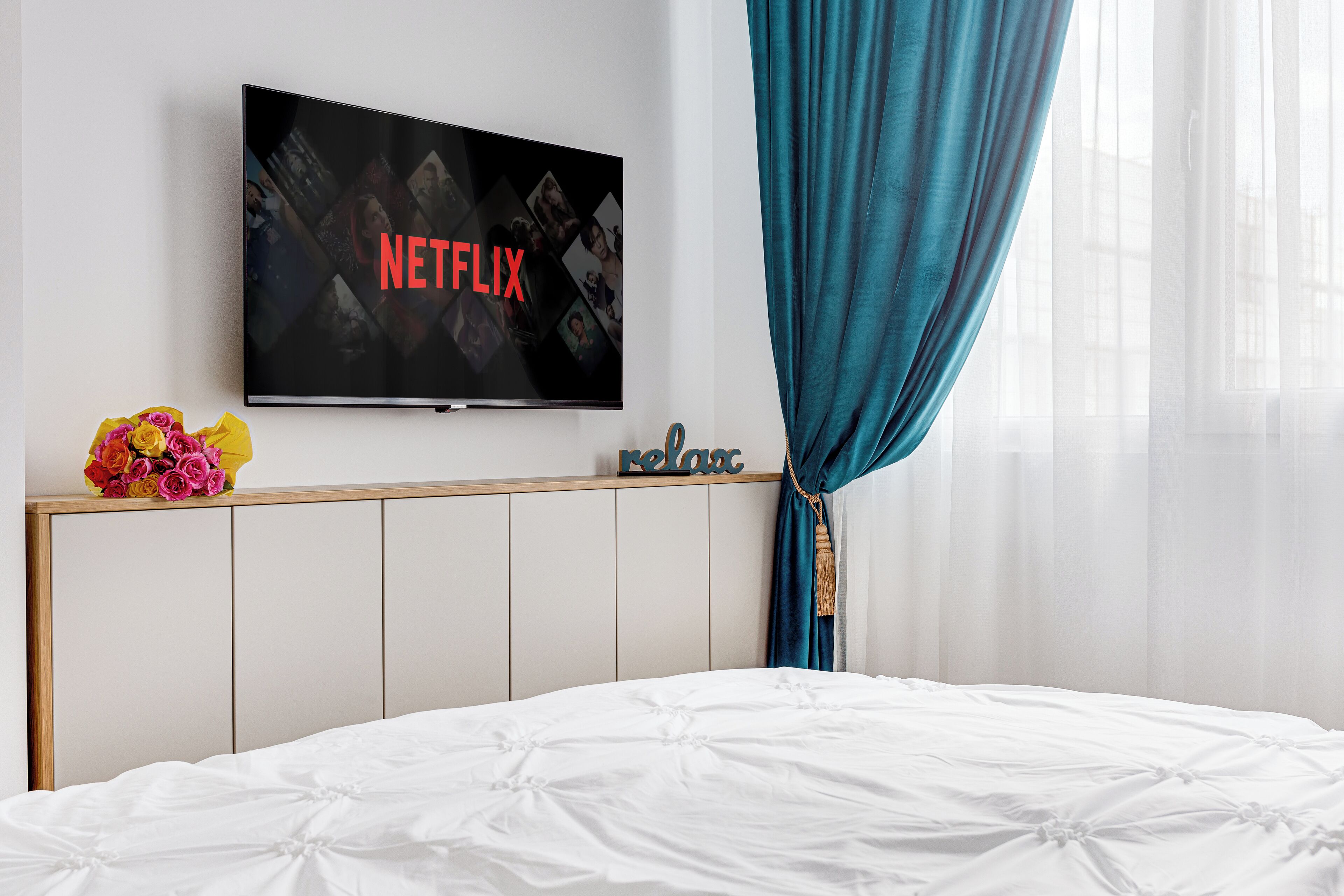 Quarto com cama, TV na parede exibindo 'NETFLIX', mesa de cabeceira com um vaso de flores e um letreiro com a palavra 'relax'.