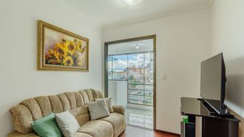 Uma sala de estar com sofá, um quadro, televisão e uma varanda com vista para a cidade.