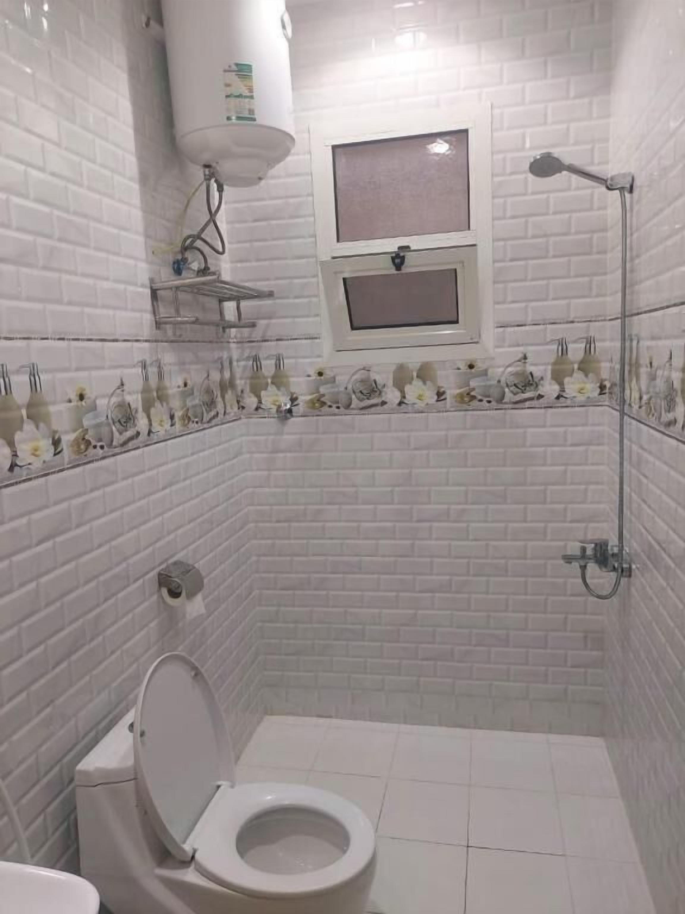 Casa de banho com azulejos brancos, sanita, chuveiro e uma pequena janela acima do espelho.