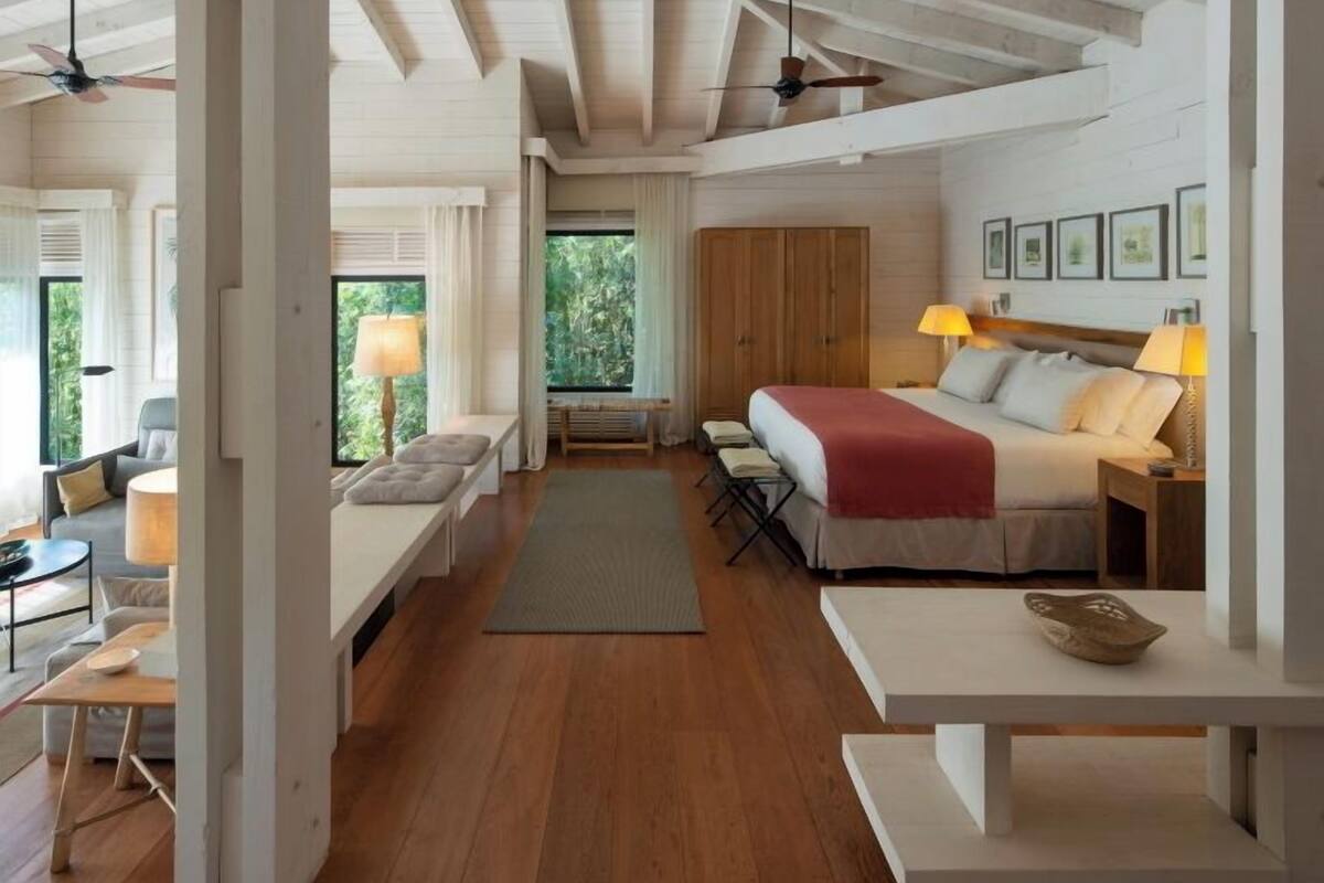 Quarto de hotel moderno com teto de madeira, uma cama com manta vermelha, uma escrivaninha e um espaço de estar com um abajur.