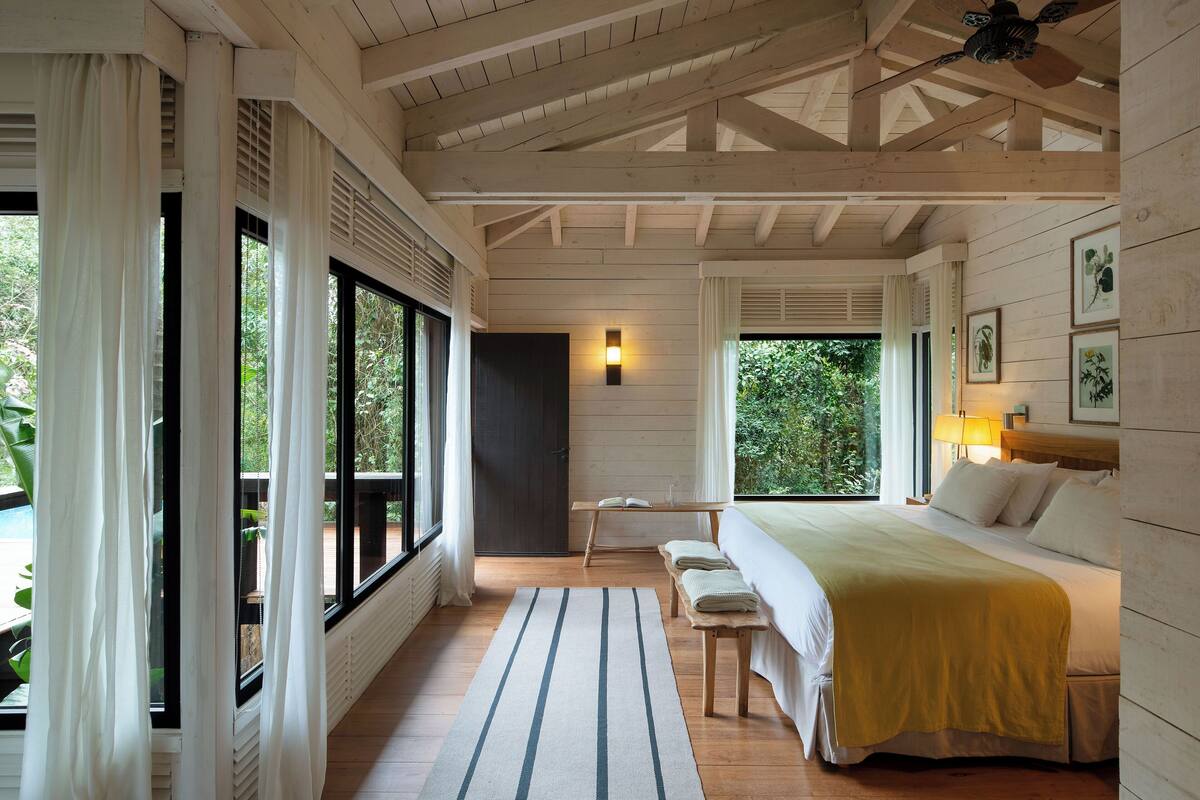 Quarto com cama grande, mesa de cabeceira, um banco e janela com cortinas.