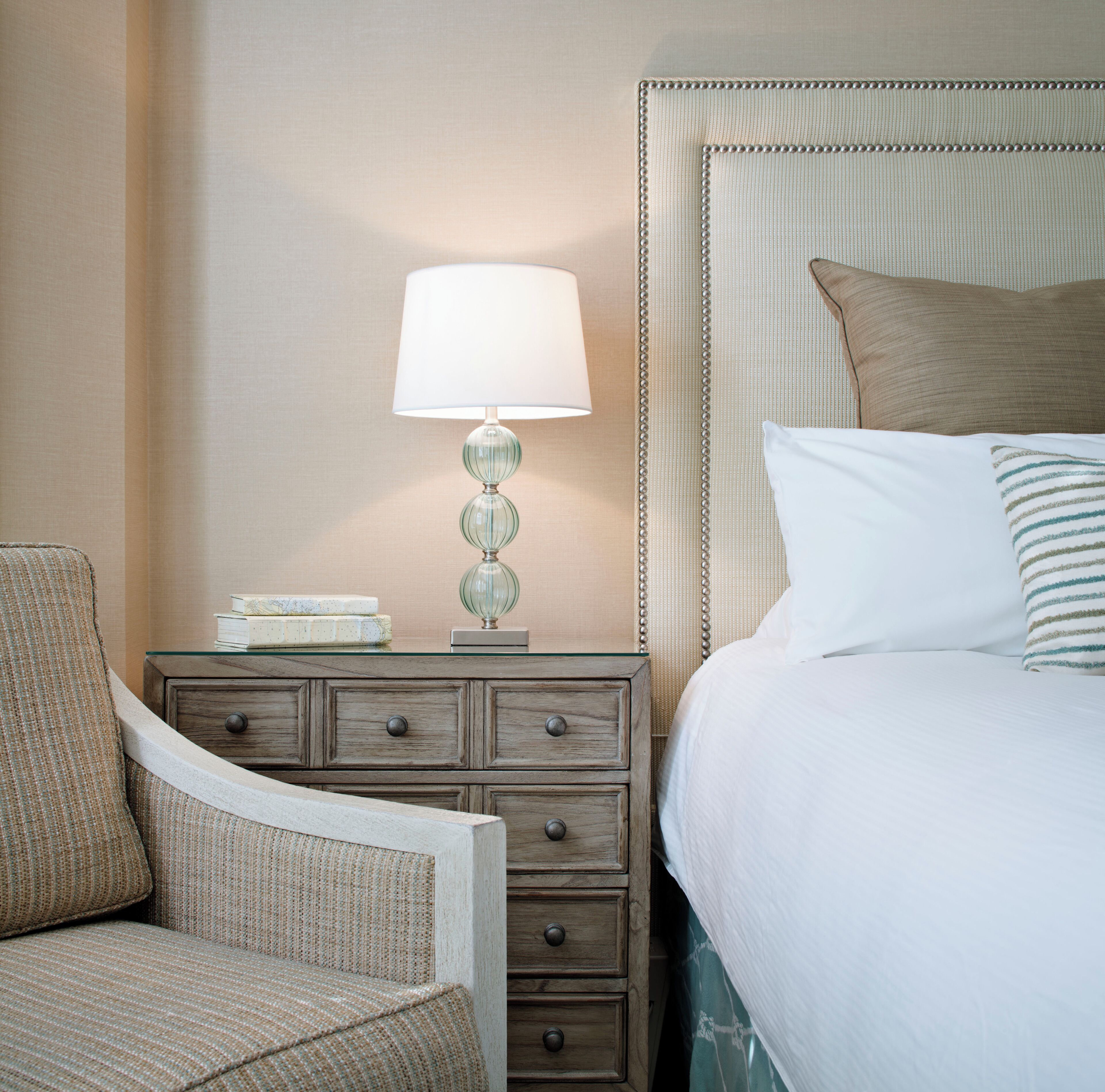 Quarto de hotel bem arrumado, com cama, mesa de cabeceira com abajur e uma cadeira.