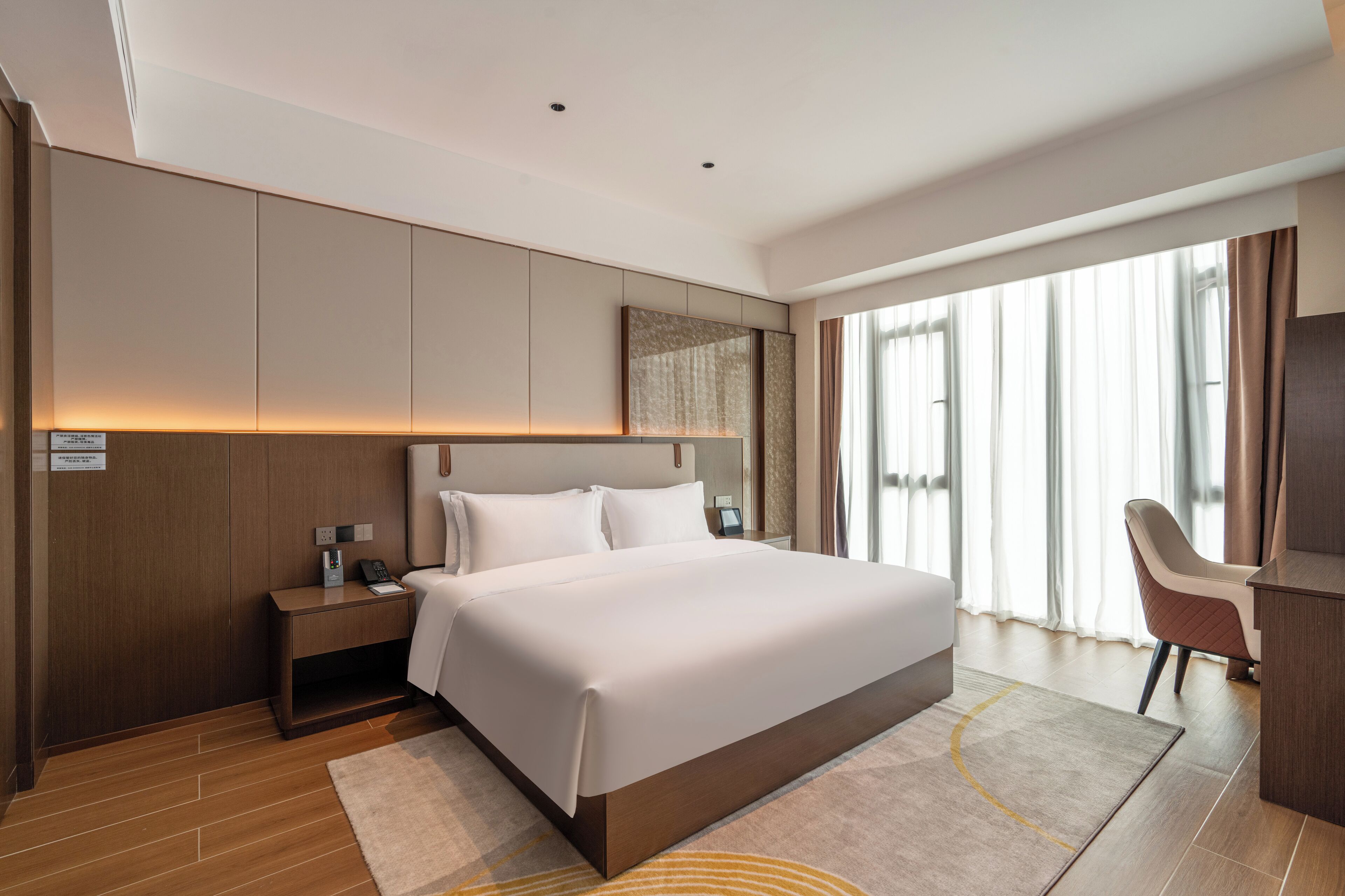 Quarto de hotel moderno com uma cama grande, mesa de cabeceira, uma cadeira e uma janela ampla com cortinas.