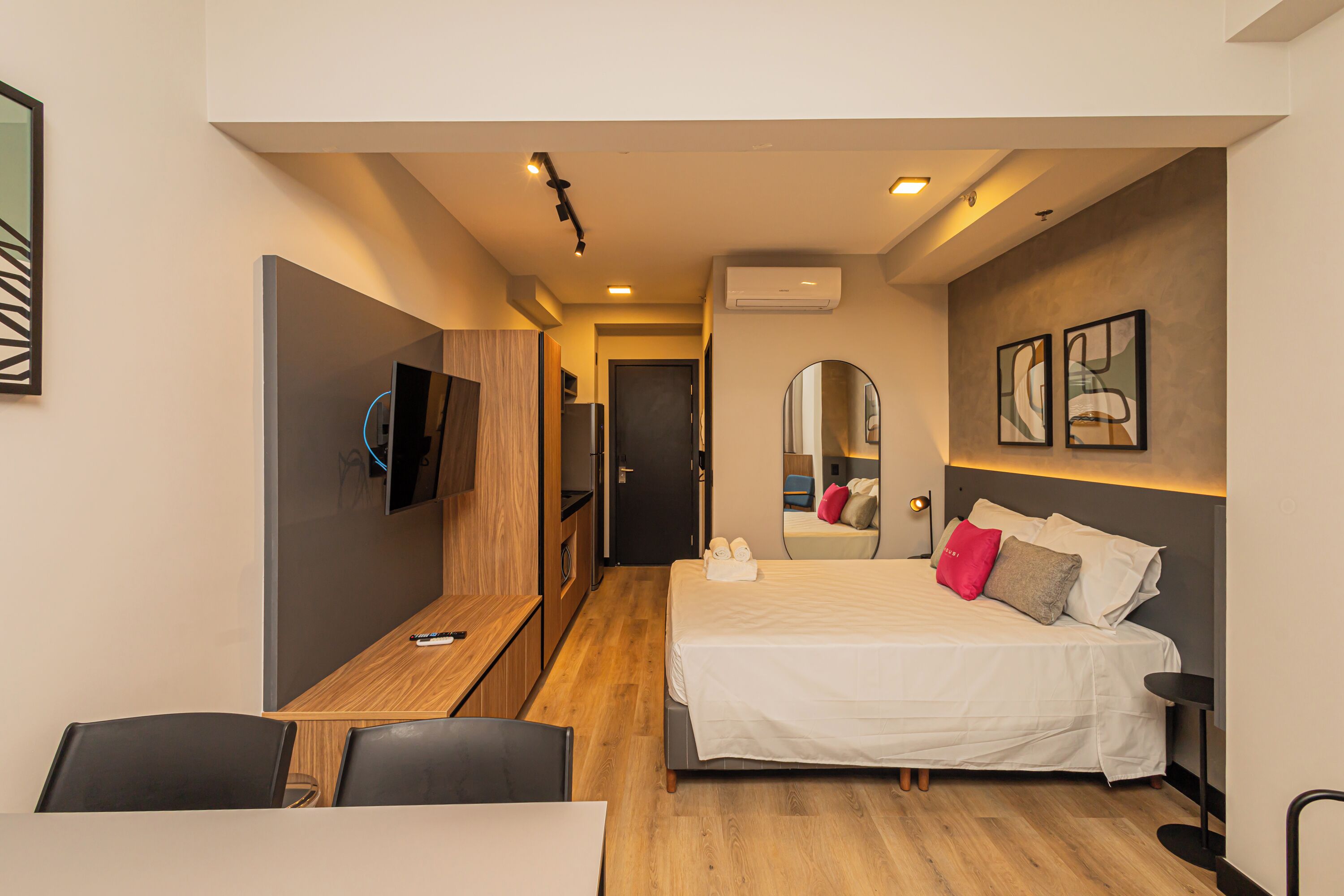 Quarto de hotel moderno com escrivaninha de madeira, cama com roupa de cama branca e várias almofadas, televisão de tela plana na parede e um espelho grande sobre a cama.