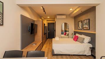 Quarto de hotel moderno com escrivaninha de madeira, cama com roupa de cama branca e várias almofadas, televisão de tela plana na parede e um espelho grande sobre a cama.