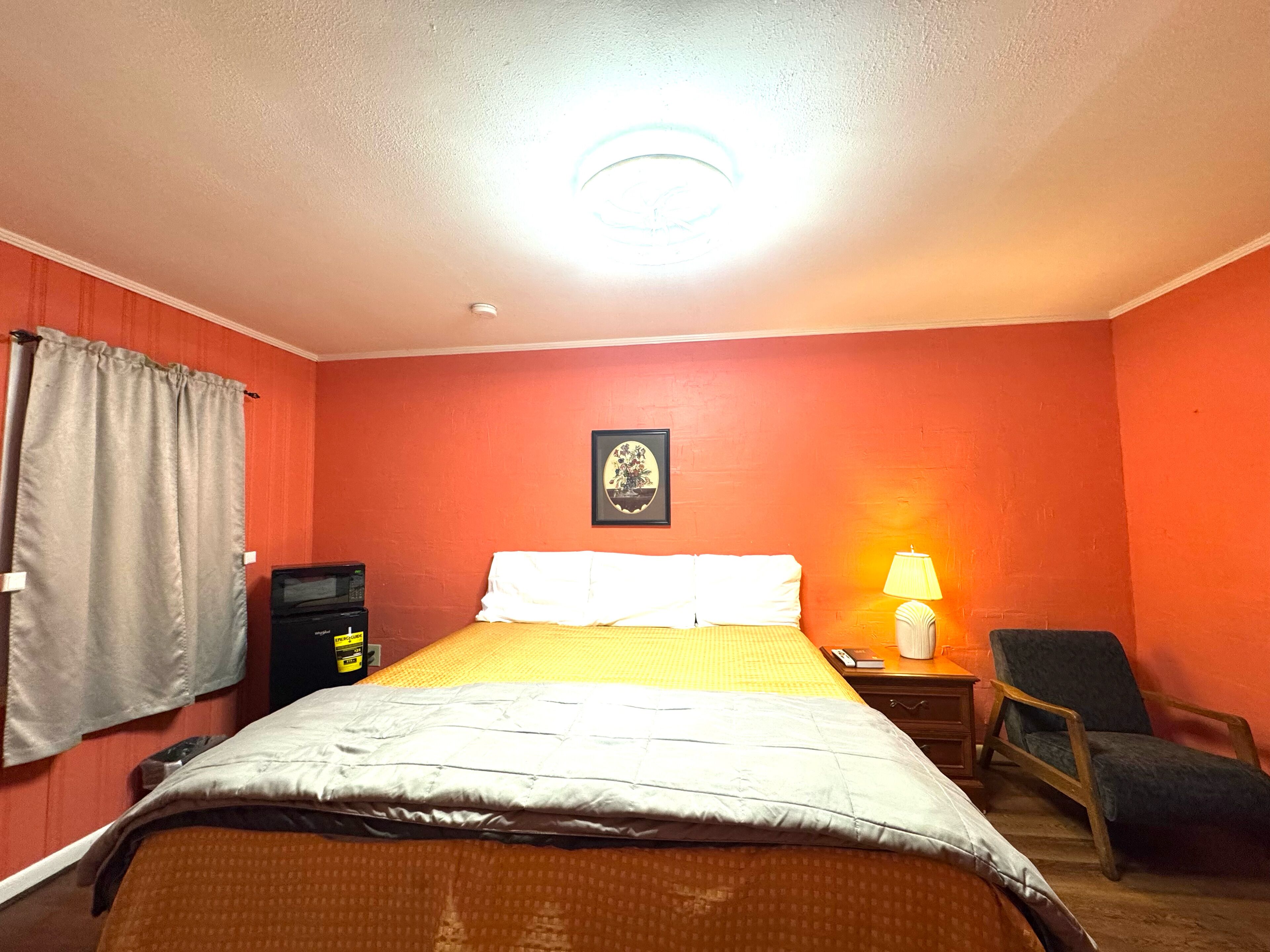 Quarto de hotel com cama, uma cadeira, mesa de cabeceira com abajur, quadro na parede e janela com cortinas.