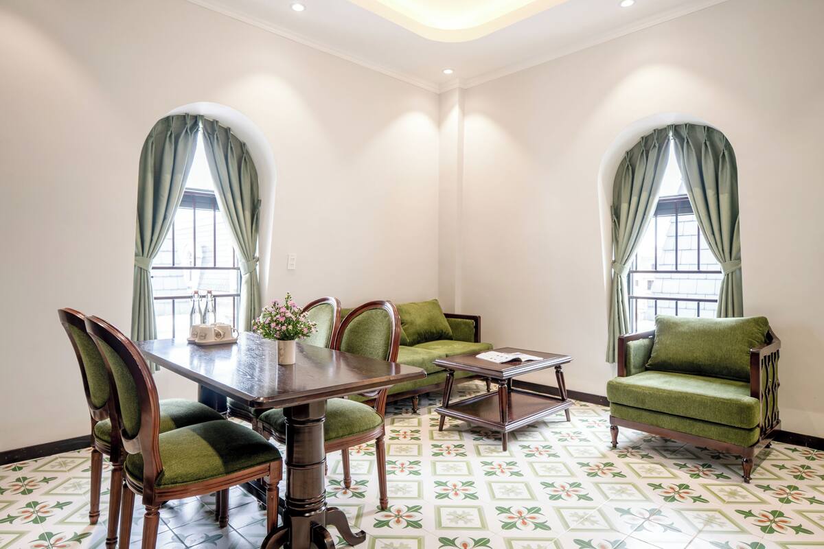 Quarto com mesa de jantar, cadeiras, sofá, mesinha de centro e duas janelas com cortinas.