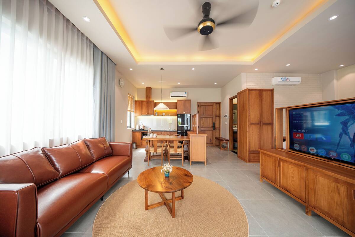 Uma sala de estar moderna com um sofá de couro, uma mesa de centro de madeira, uma televisão e uma área de cozinha ao fundo.