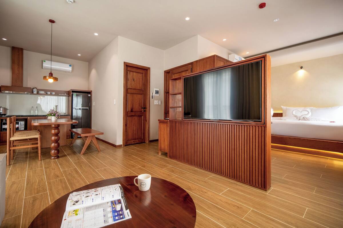 Quarto de hotel moderno com piso de madeira, área de jantar com mesa e cadeiras, cozinha com refrigerador e uma cama grande com televisão.