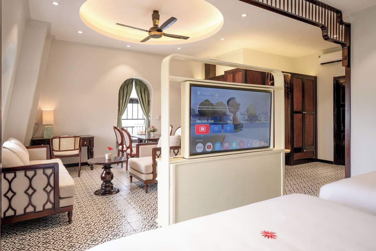 Quarto de hotel com uma televisão de tela grande, uma cama, um sofá, área de jantar com cadeiras e um lance de escadas.