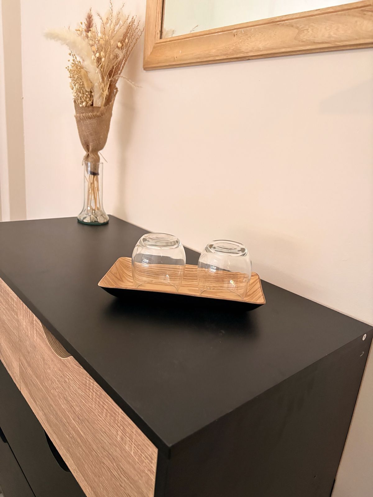 Uma mesa preta com um tabuleiro de madeira que contém dois copos de vidro, um vaso com flores secas e um espelho na parede.