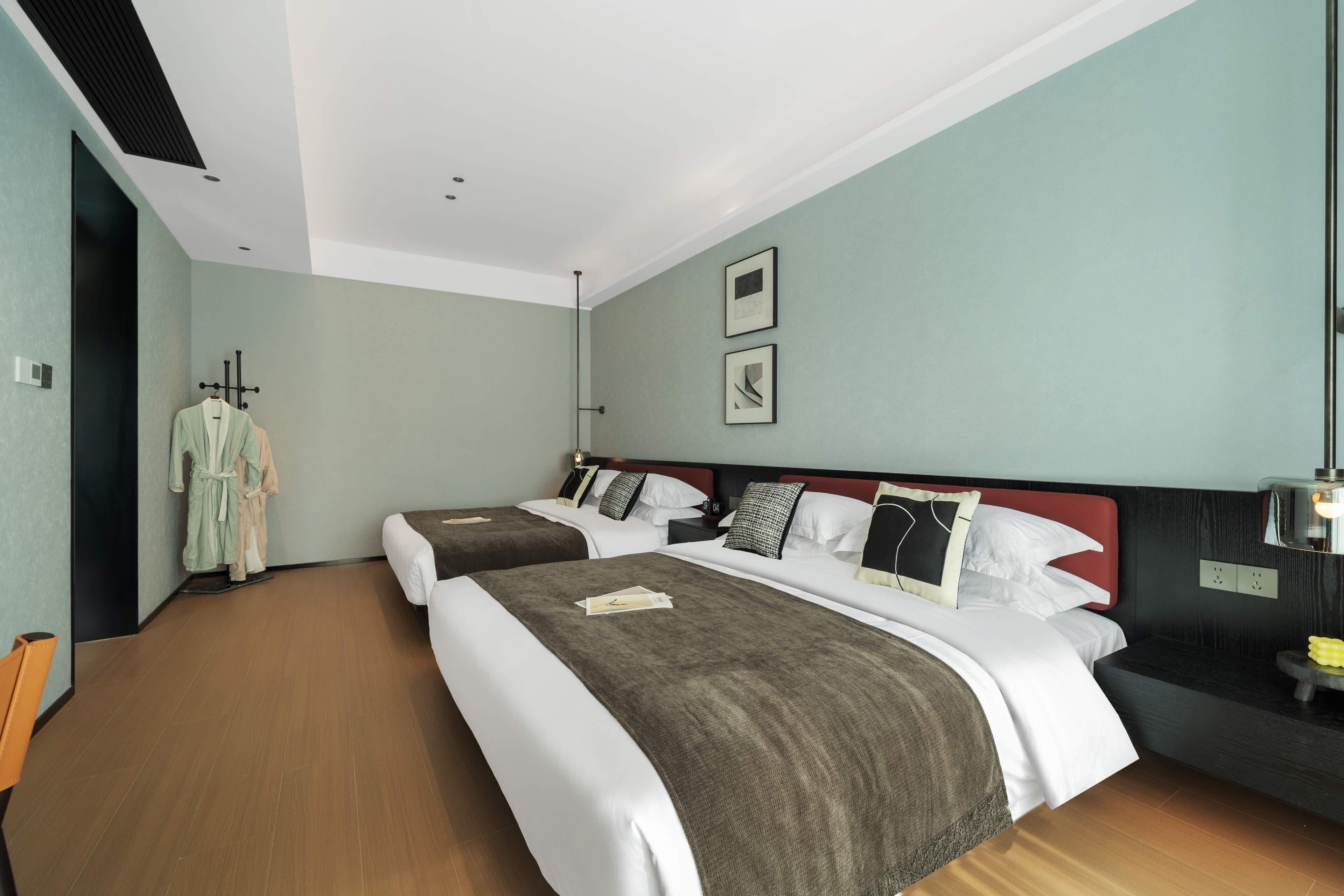 Quarto de hotel com duas camas, piso de madeira, cabideiro e mesa de cabeceira com abajur.