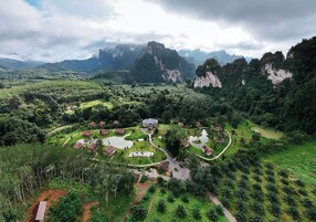 Um resort com vários edifícios tipo bangalô, uma piscina central e uma paisagem exuberante circundante, com montanhas ao fundo.