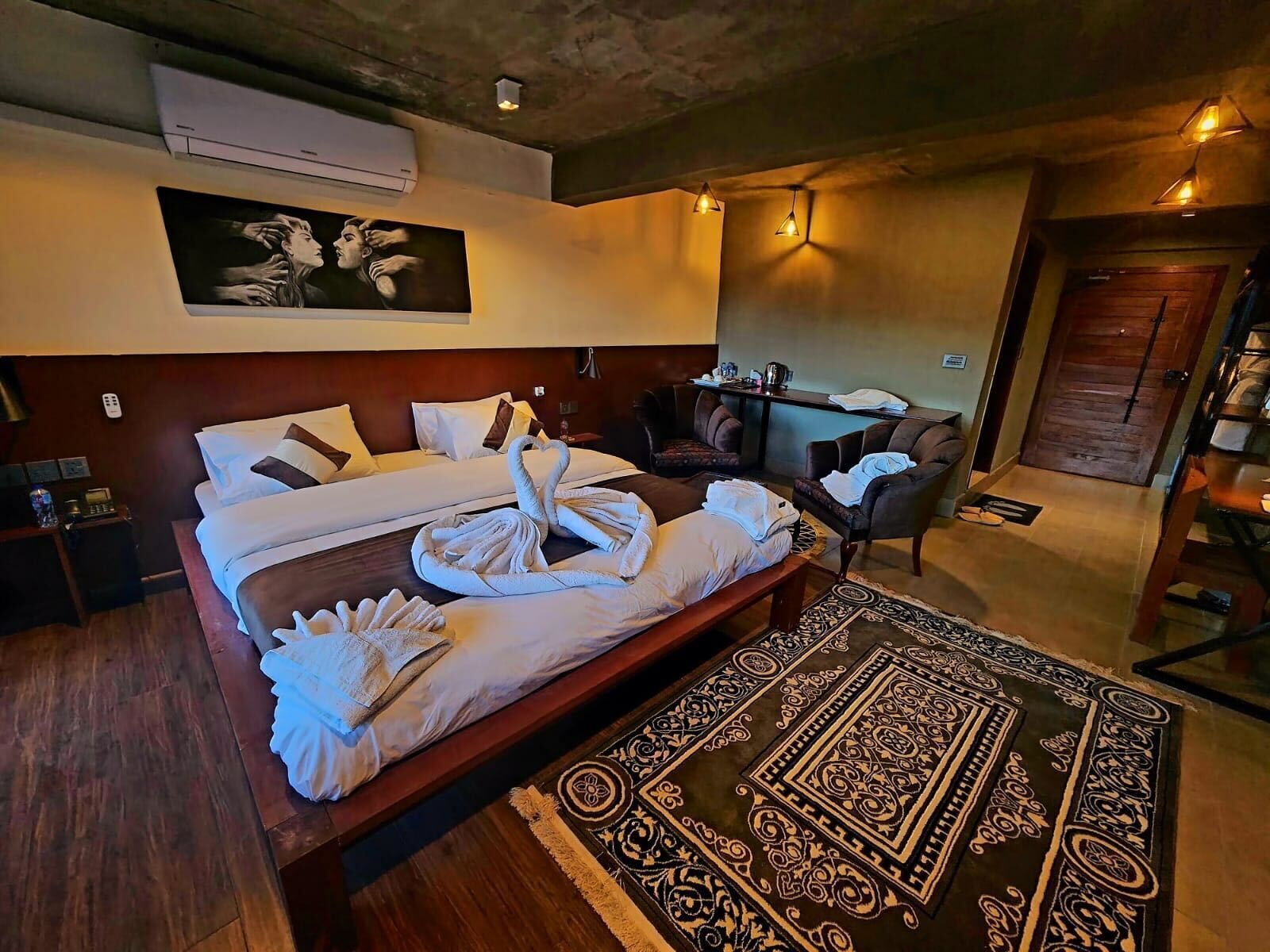 Quarto de hotel com uma cama grande, uma escrivaninha e uma cadeira. Há um quadro na parede e uma porta ao fundo.